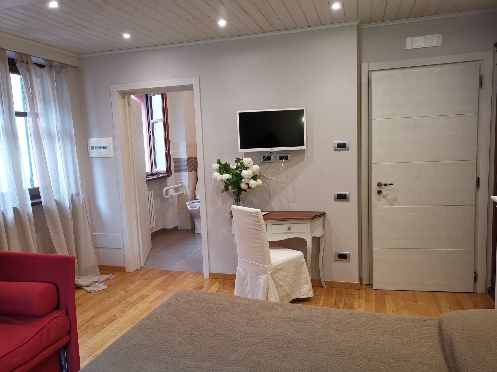 Uma casa de banho moderna com sanita, chuveiro e lavatório. Há uma televisão na parede e uma porta com puxador.