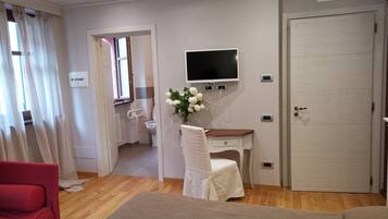 Uma casa de banho moderna com sanita, chuveiro e lavatório. Há uma televisão na parede e uma porta com puxador.