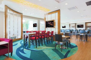 Um hall de hotel moderno com uma ampla área de estar, várias telas de TV e um tapete colorido.
