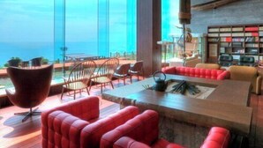 Interior moderno com uma grande janela com vista para o mar, assentos estofados em vermelho e uma estante de livros.