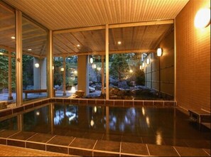 Piscina ampla em ambiente interno com paredes de vidro transparente, teto de madeira e piso revestido, refletindo as árvores e luzes ao redor.