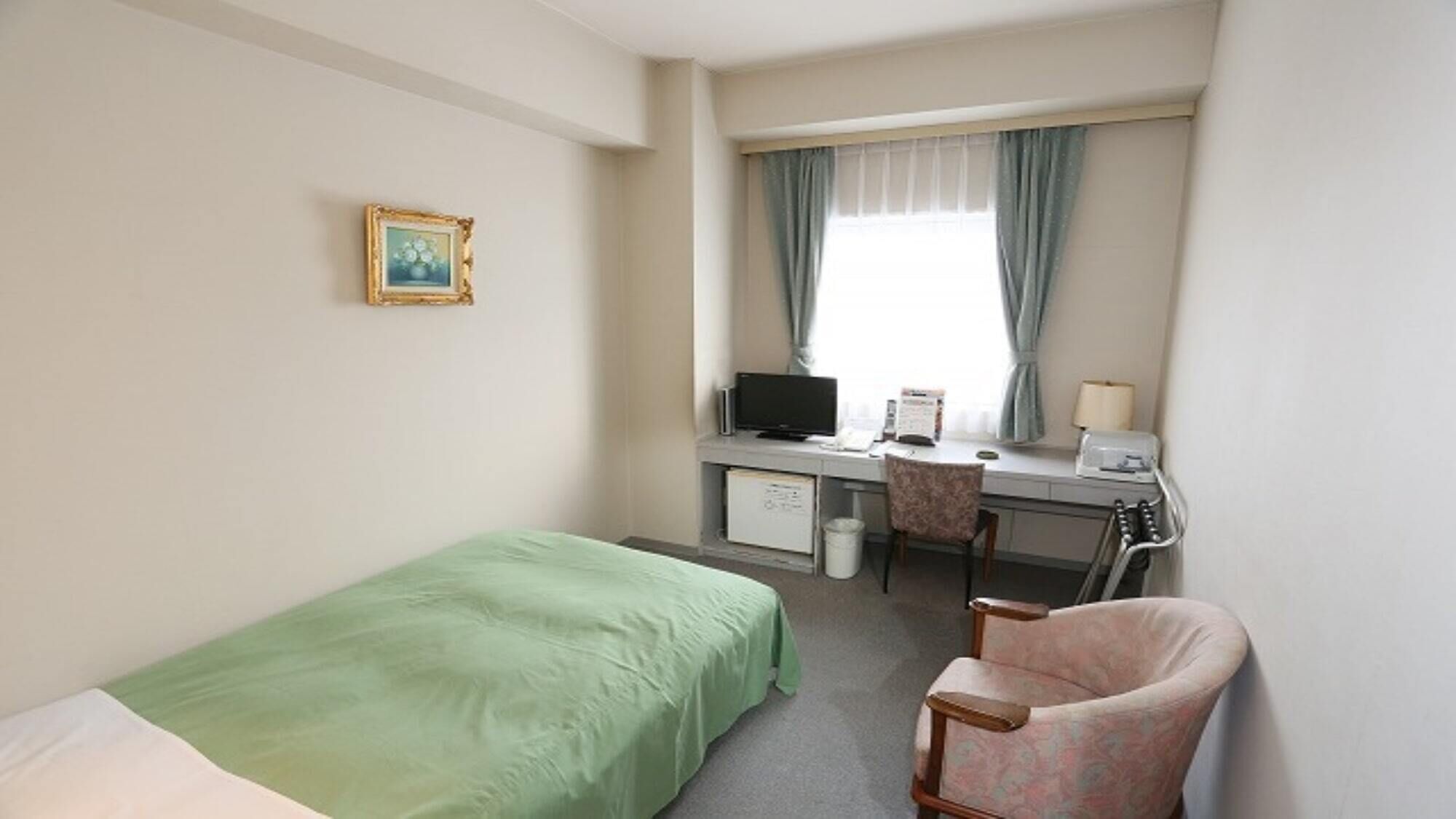Quarto de hotel com uma cama de solteiro, escrivaninha com computador, cadeira e janela com cortinas.