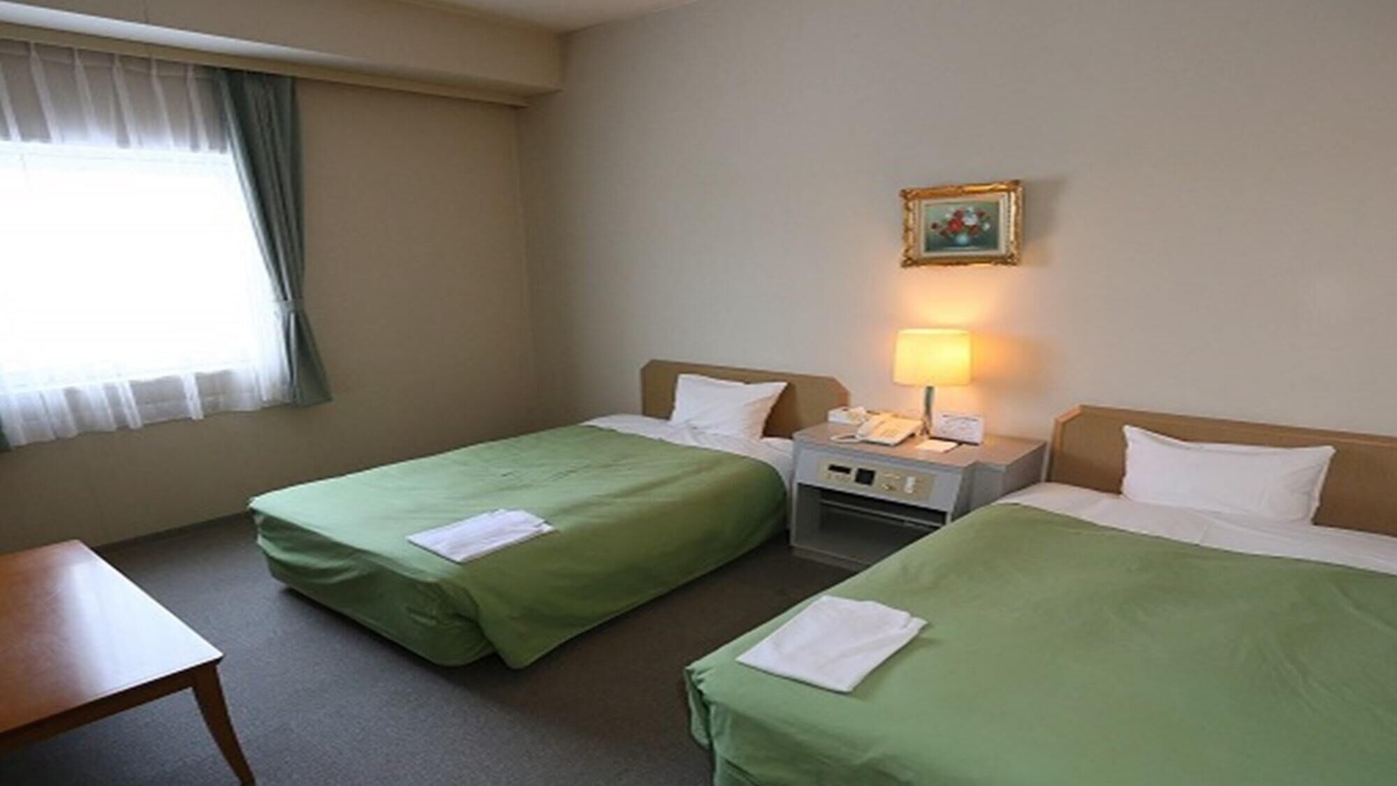 Quarto de hotel com duas camas de solteiro, mesa de cabeceira com abajur, uma mesinha e uma janela com cortinas.