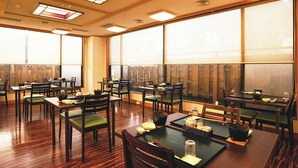 Restaurante com chão de madeira, mesas, cadeiras e janelas amplas.