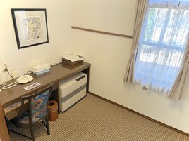 Quarto de hotel com uma secretária de madeira, um telefone, uma impressora, uma cadeira, um cesto de lixo, um quadro na parede e uma janela com cortinas.
