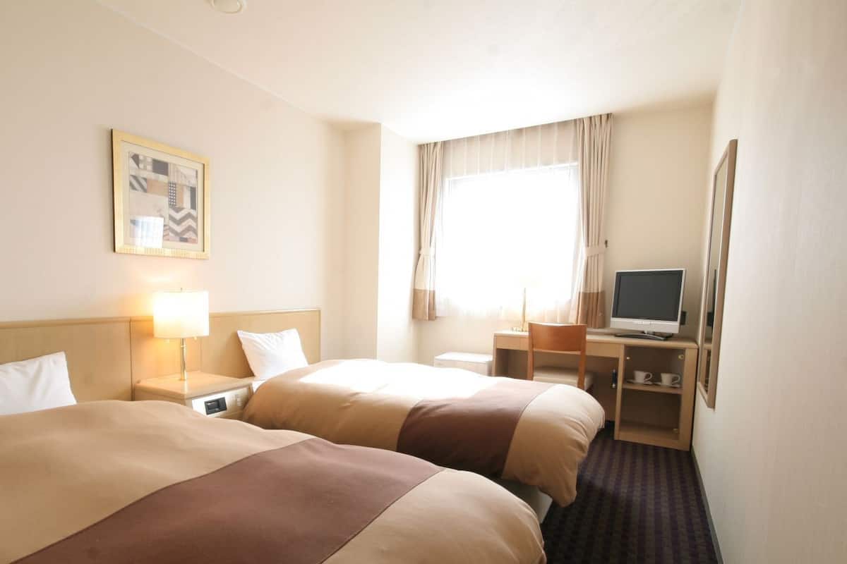 Quarto de hotel com duas camas, uma escrivaninha com computador, televisão e janela com cortinas.