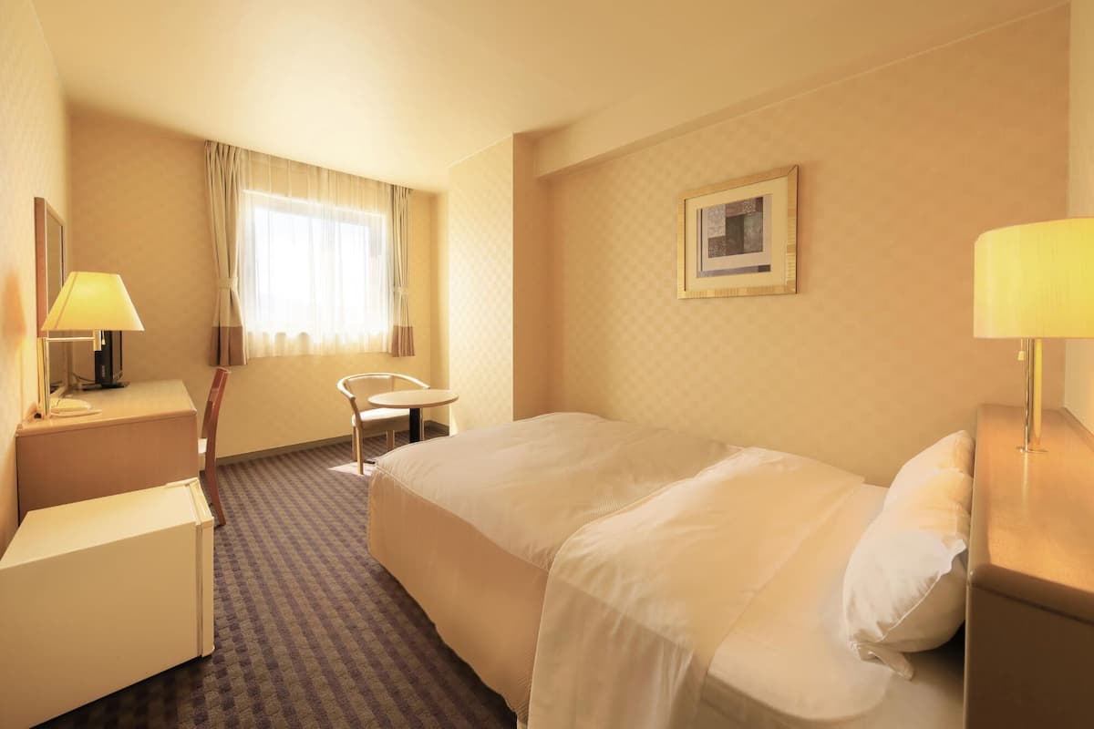 Quarto de hotel com cama, escrivaninha, cadeira, luminária e janela com cortinas.