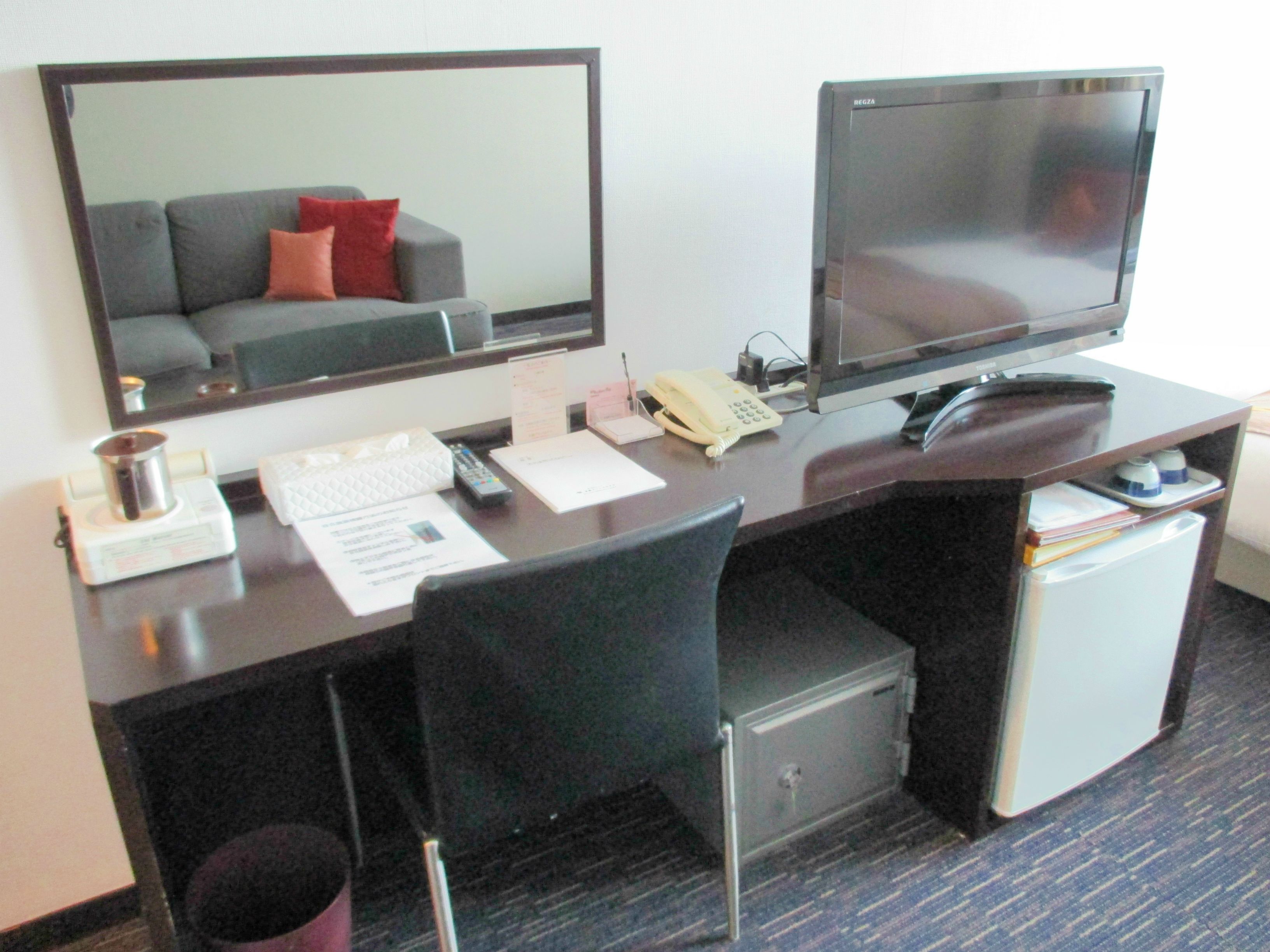 Quarto de hotel com escrivaninha, computador, telefone, cofre pequeno e espelho.