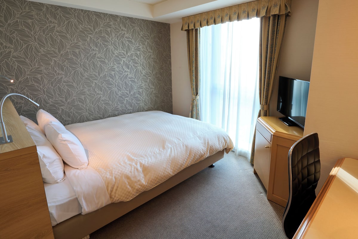 Quarto de hotel com cama, escrivaninha, cadeira, televisão e janela com cortinas.