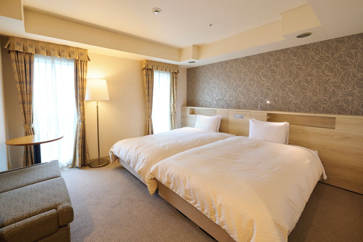 Quarto de hotel com duas camas, uma escrivaninha, uma lâmpada e parede estampada.