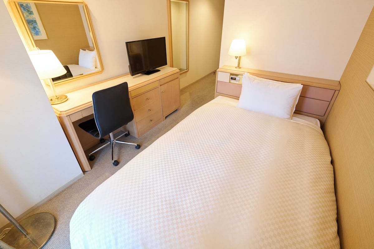 Quarto de hotel com cama, secretária com computador, cadeira, televisão e mesa de cabeceira com abajur.
