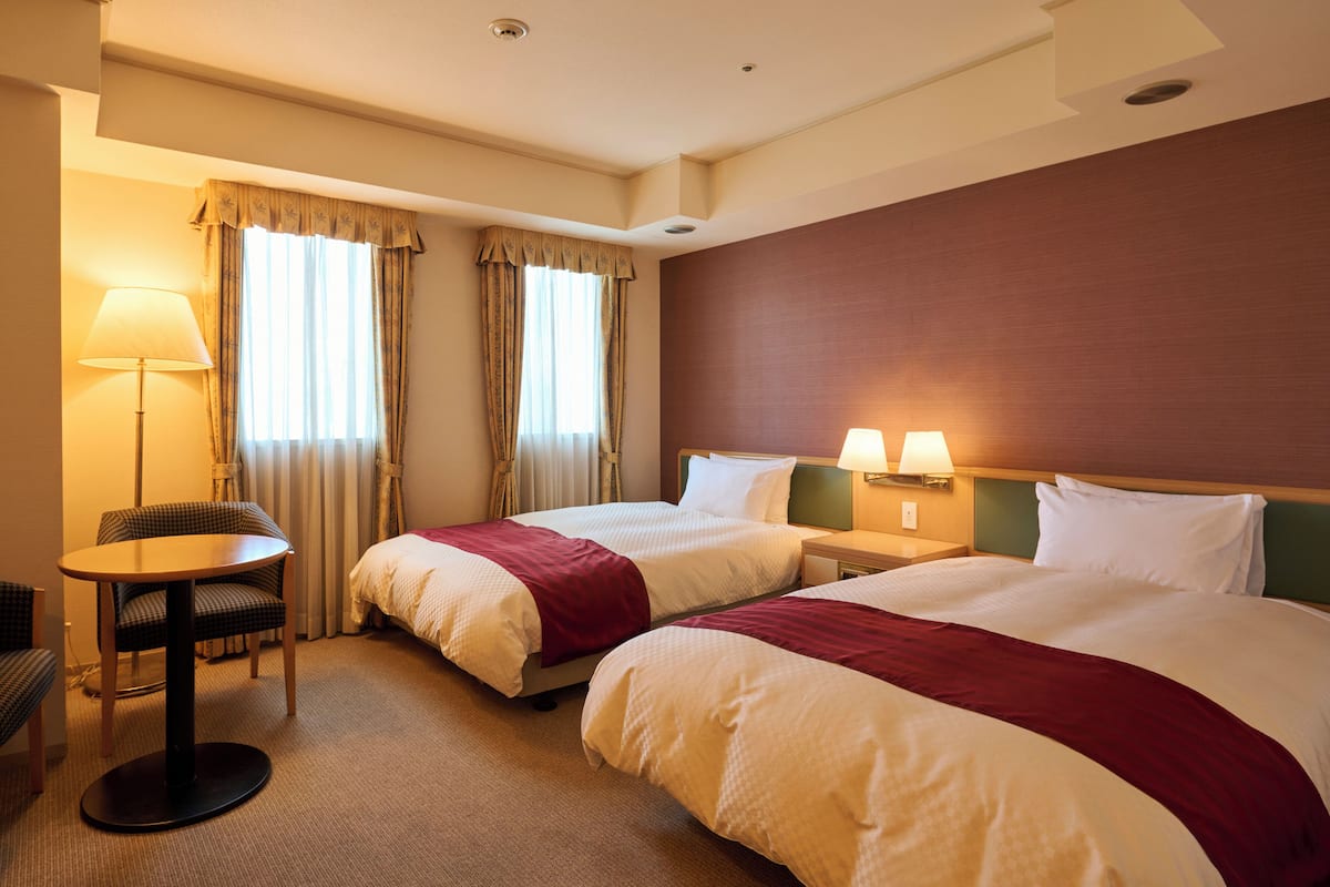 Quarto de hotel com duas camas, uma escrivaninha, uma cadeira e uma luminária.