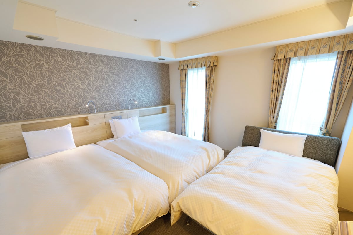 Quarto de hotel com duas camas, cabeceira estampada com folhagens e janela com cortinas.