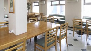 Uma sala de jantar ampla, com mesas e cadeiras de madeira, grandes janelas e um ar condicionado na parede.
