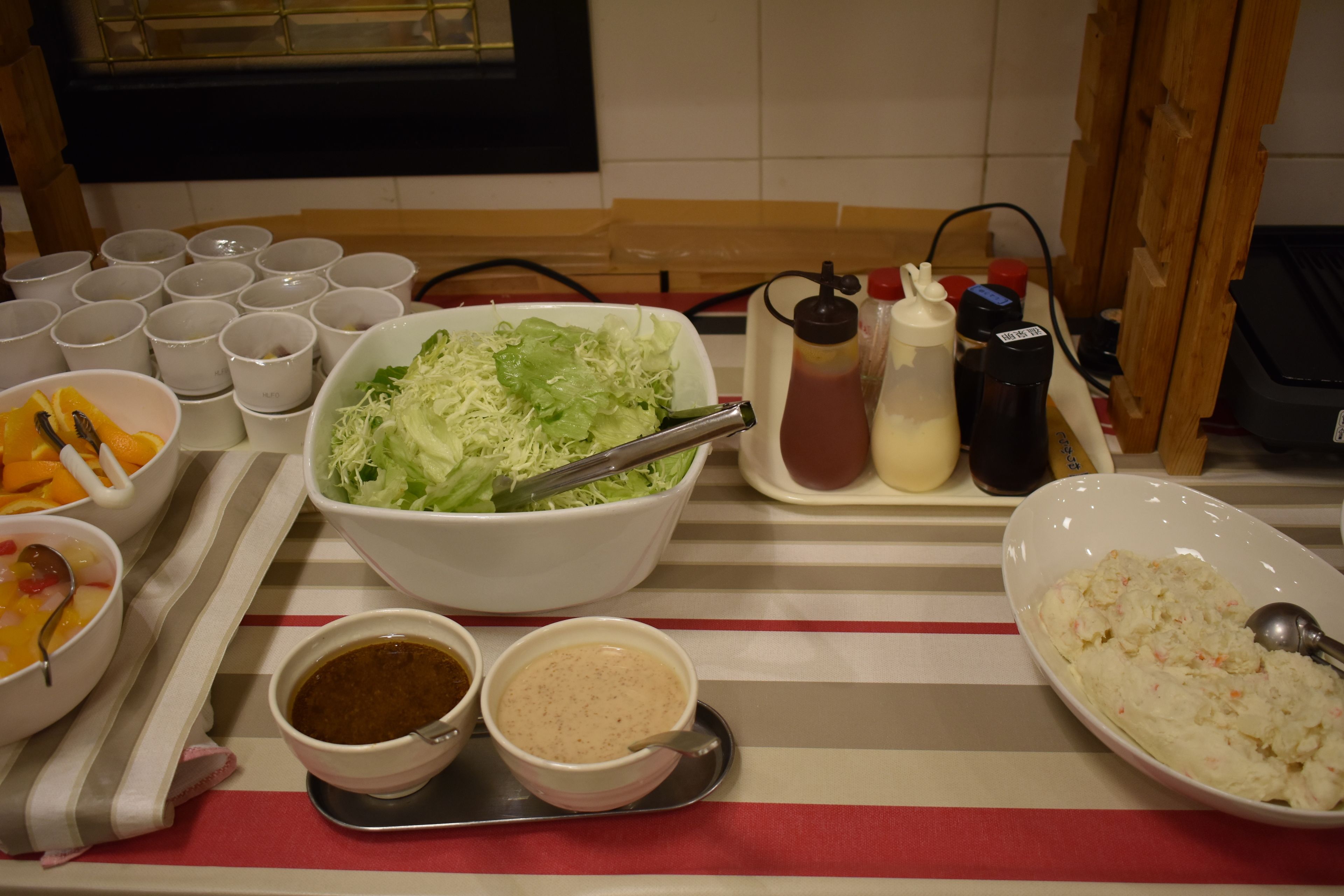 Mesa de buffet com diversos pratos, incluindo salada, sopa e arroz, além de acompanhamentos e copos.