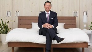 Um homem sentado em uma cama num quarto de hotel, com cabeceira de madeira, roupa de cama branca e plantas em vasos.