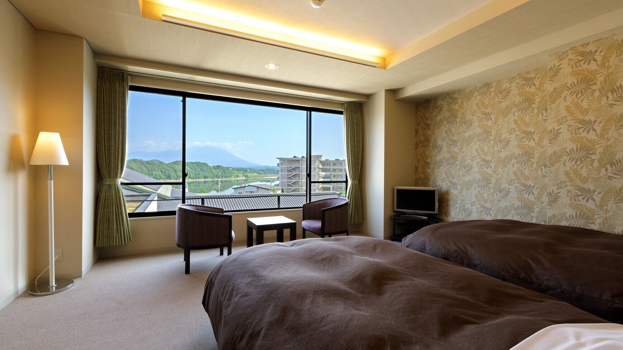 Quarto de hotel com duas camas, uma escrivaninha, uma cadeira, televisão e uma janela ampla com vista para a cidade e montanhas.