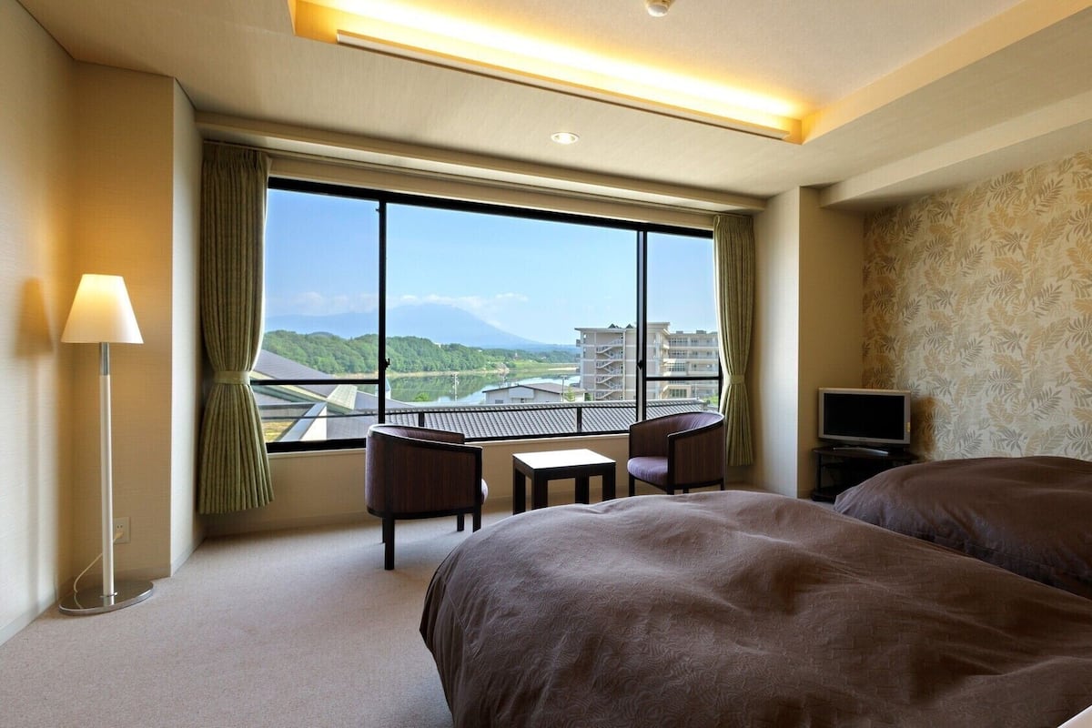 Quarto de hotel com duas camas, uma escrivaninha, uma cadeira, televisão e uma janela ampla com vista para a cidade e montanhas.