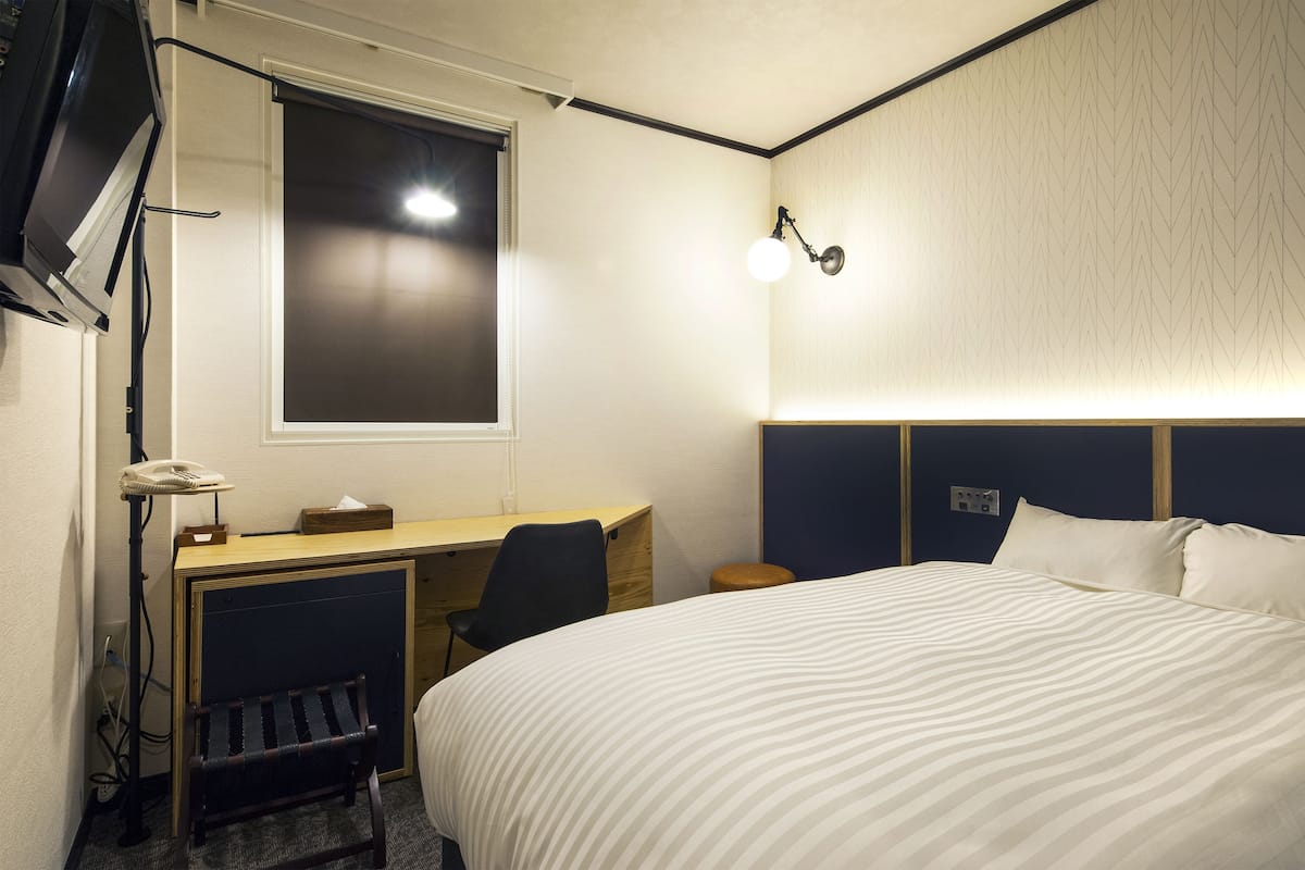Quarto de hotel com uma cama, uma escrivaninha, uma cadeira, um abajur e uma janela.