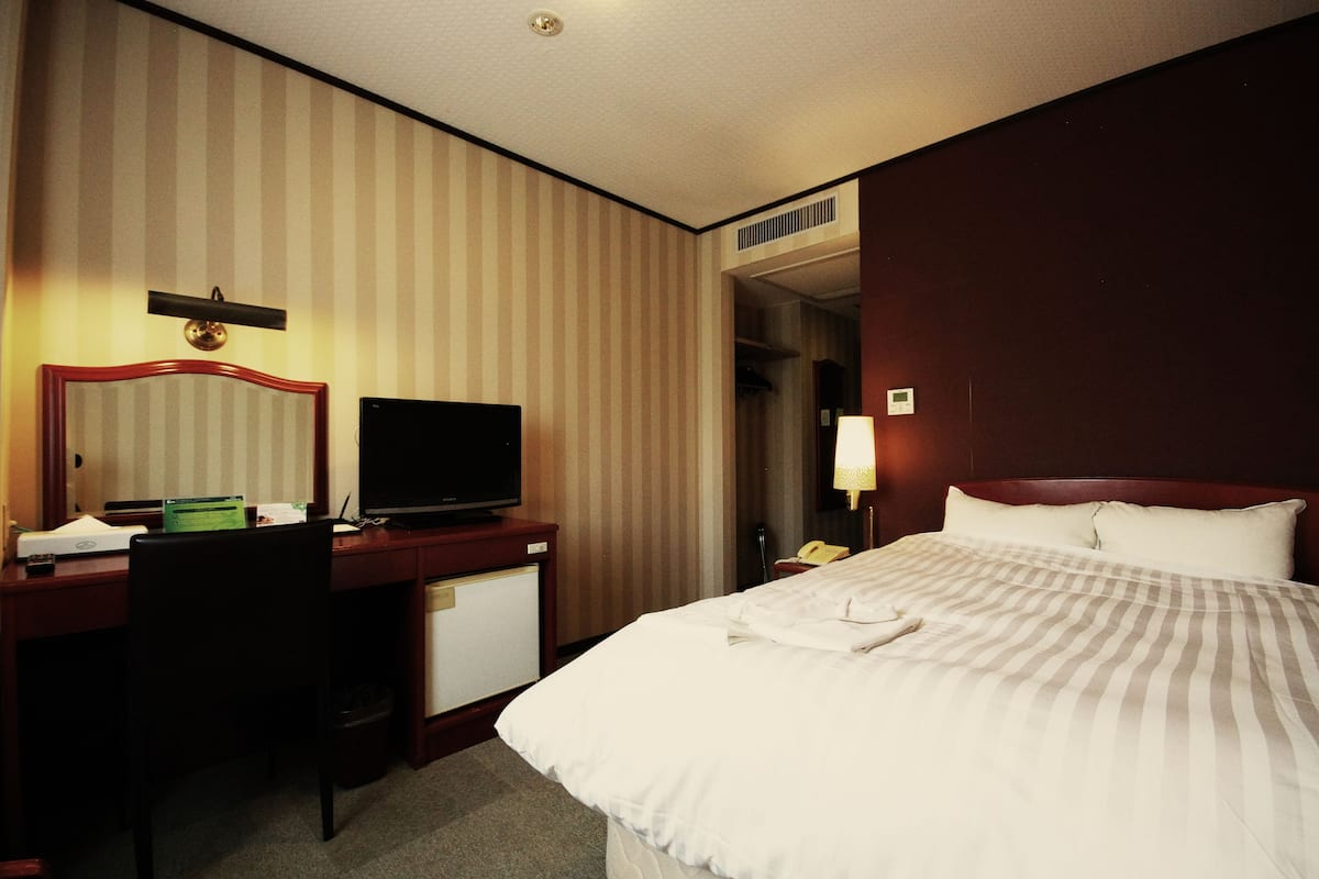 Quarto de hotel com cama, escrivaninha com computador, televisão e luminária.