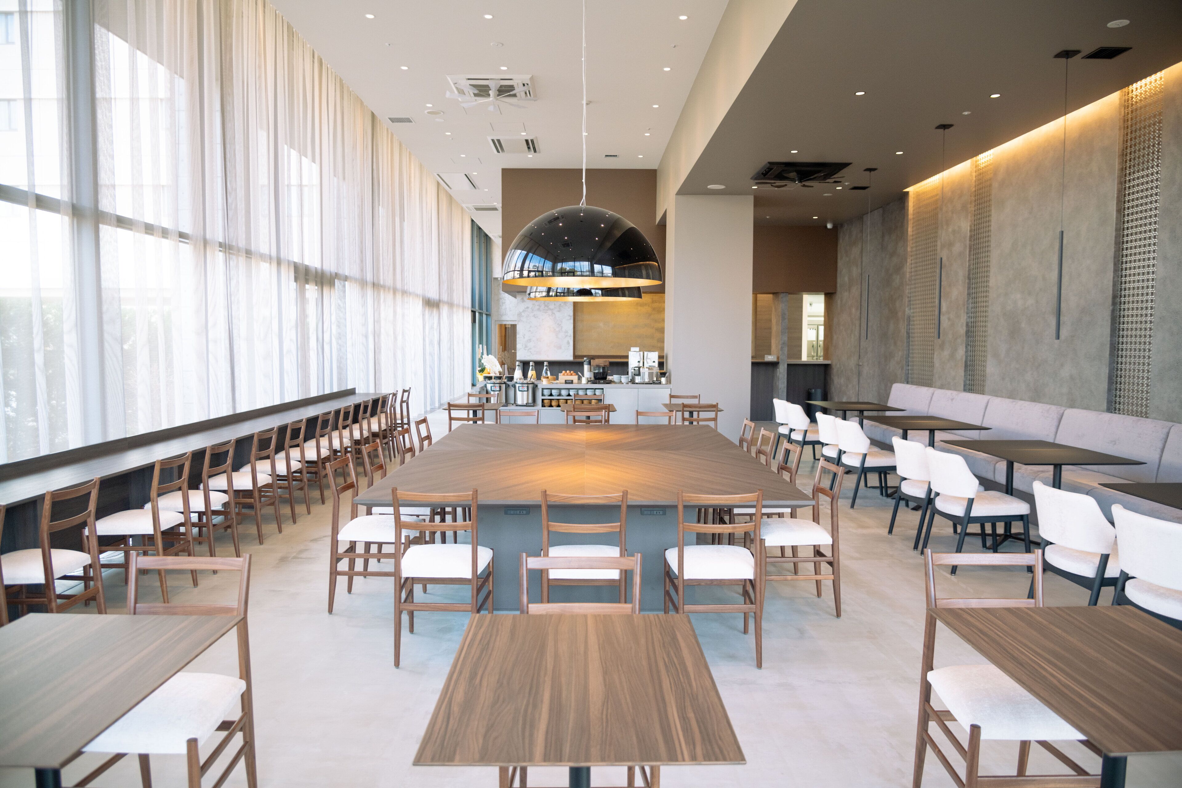 Uma sala de jantar espaçosa, com mesas e cadeiras compridas, uma área de cozinha central e grandes janelas que proporcionam iluminação natural.