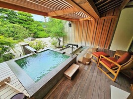 Uma banheira de hidromassagem com deck de madeira, rodeada por vegetação e um edifício em estilo japonês tradicional.
