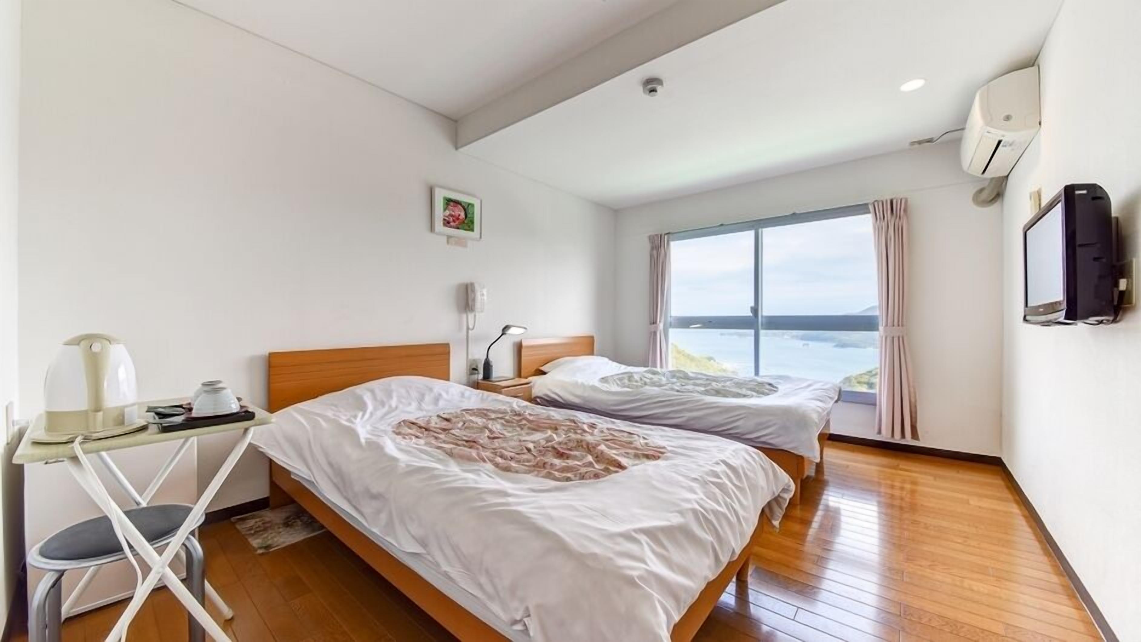 Quarto de hotel com duas camas, uma escrivaninha com chaleira, uma mesinha de cabeceira, uma janela com vista e ar condicionado.