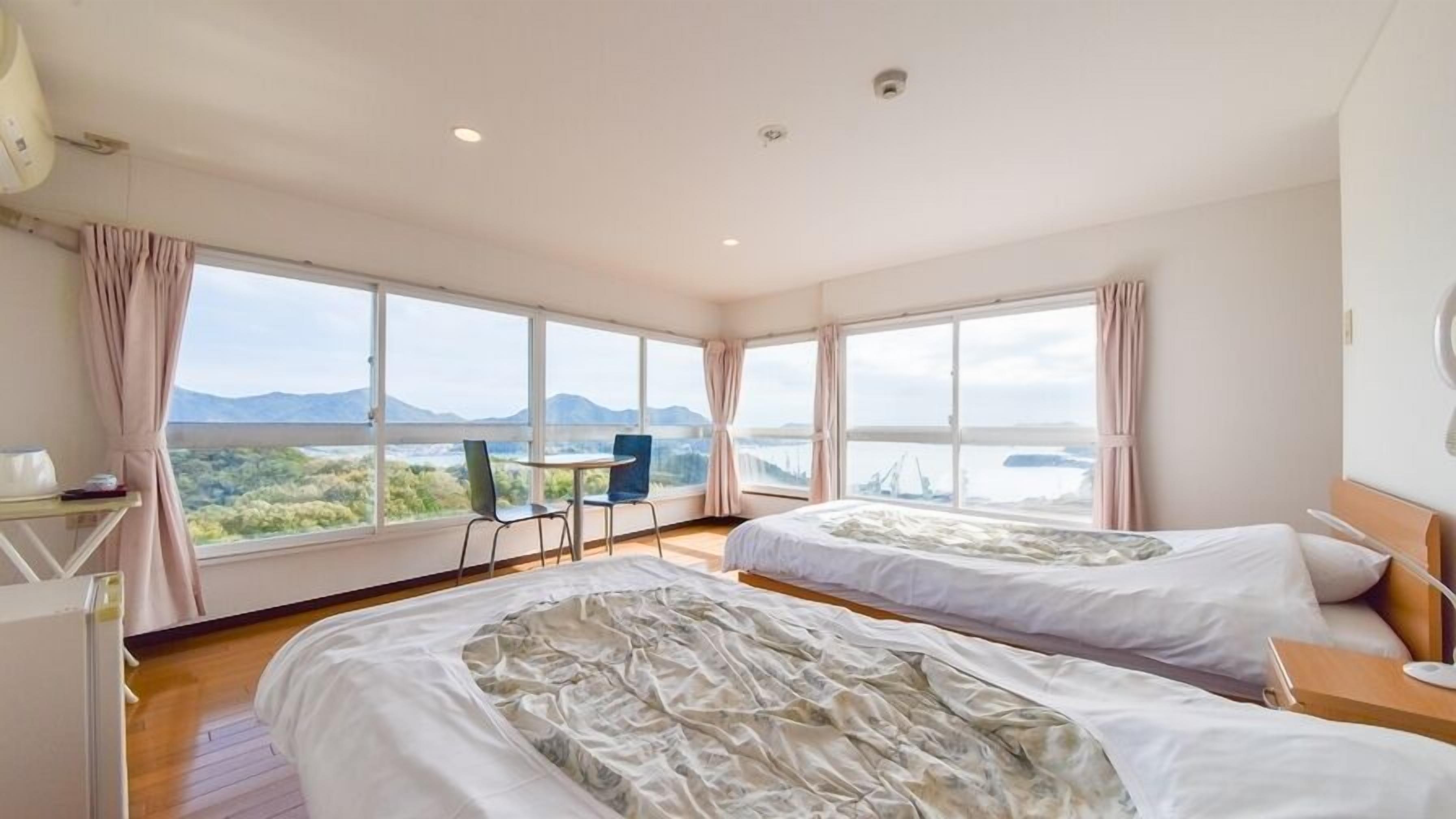 Quarto de hotel com duas camas, uma escrivaninha, uma cadeira e grandes janelas com vista para a paisagem.