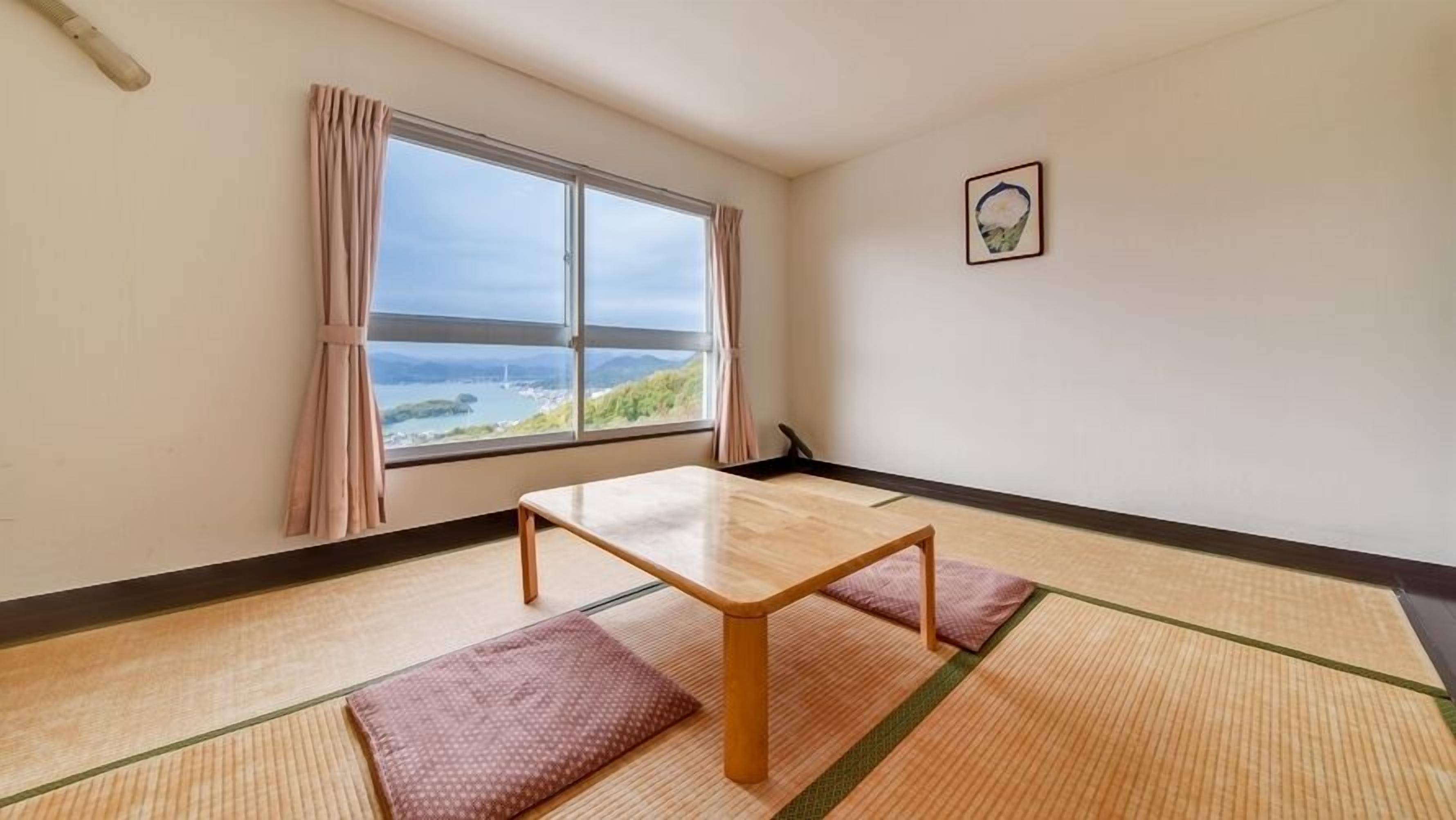 Quarto com piso de tatame, uma mesa de madeira e vista para a paisagem costeira através de uma grande janela.