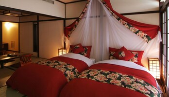 Quarto japonês tradicional com cama de dossel, roupa de cama com padrão floral vermelho e vista para um espaço japonês tradicional com tatames e uma mesa baixa.