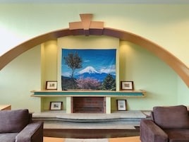 Uma lareira com um quadro emoldurado acima, ladeada por duas poltronas, e um grande tapeçaria retratando uma paisagem de montanha.
