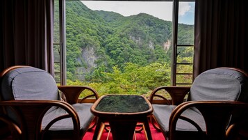 Quarto com duas cadeiras de vime e uma pequena mesa, com vista para um vale verdejante e montanhas.