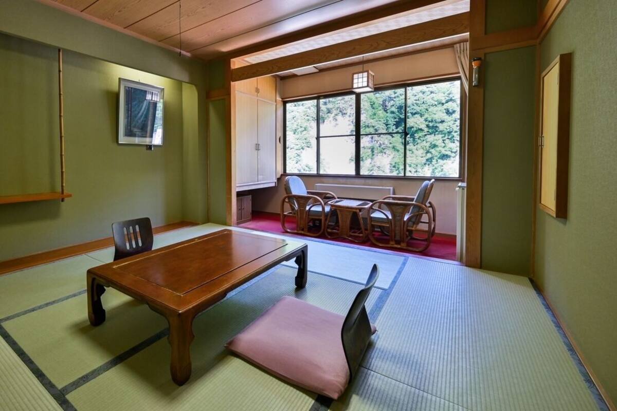 Um quarto tradicional com tatames, uma mesa de madeira, janelas com vista para árvores e teto de madeira baixo.