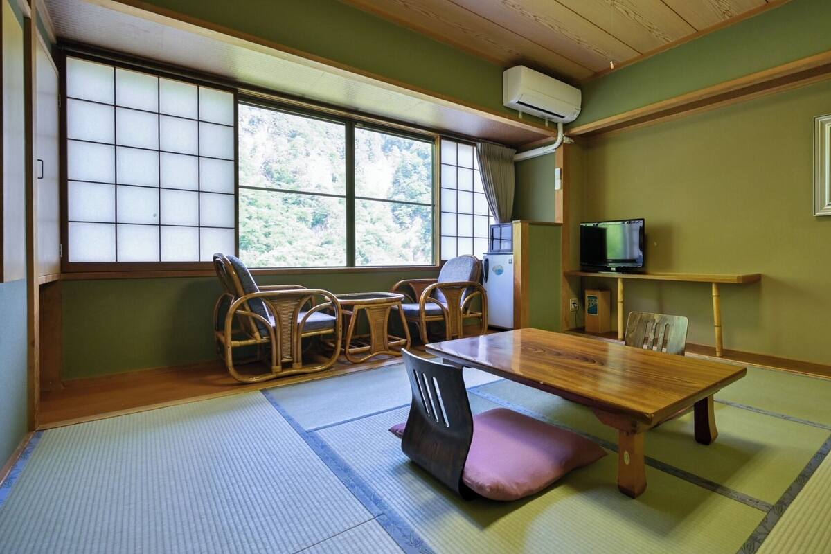 Um quarto com tatames, uma mesa de madeira, cadeiras, televisão e uma janela com vista para árvores.