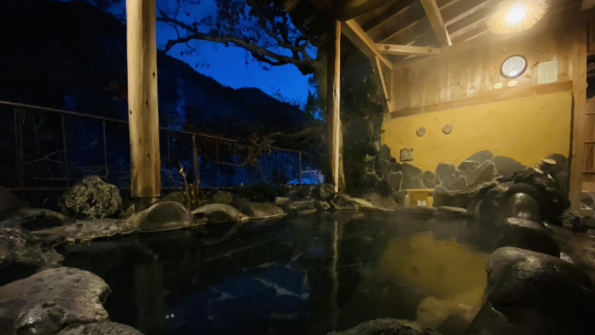 Um onsen japonês tradicional com estrutura de madeira, banheira de pedra e vista para as montanhas ao entardecer.