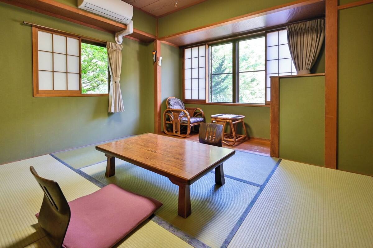 Um espaço com tatames, uma mesa de madeira, cadeiras e vista para o verde lá fora.
