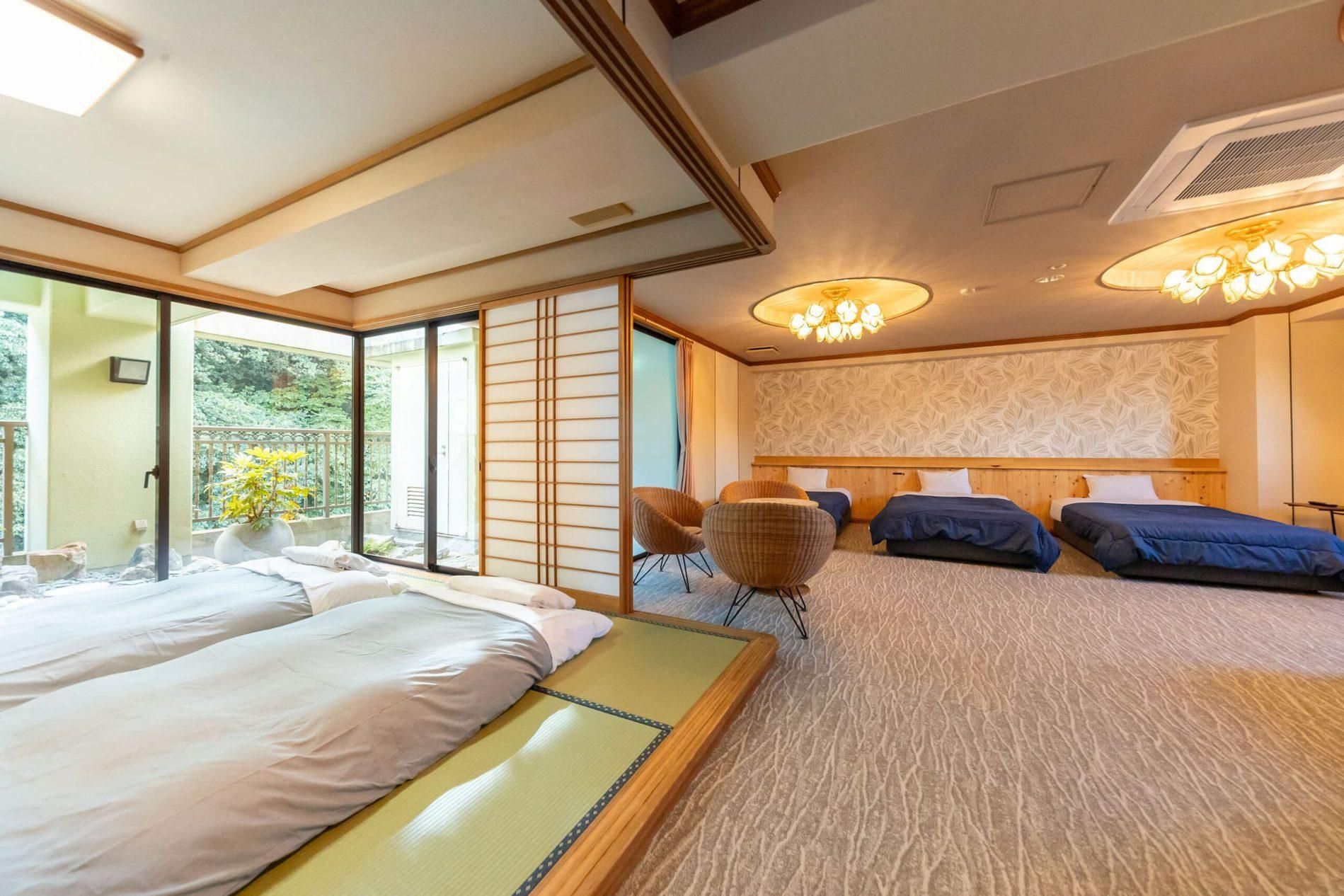 Quarto de hotel com cama, duas mesas de cabeceira, uma cadeira, um sofá e vista para área verde pelas janelas.