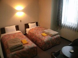 Quarto de hotel com duas camas de solteiro, uma mesinha e uma cadeira.