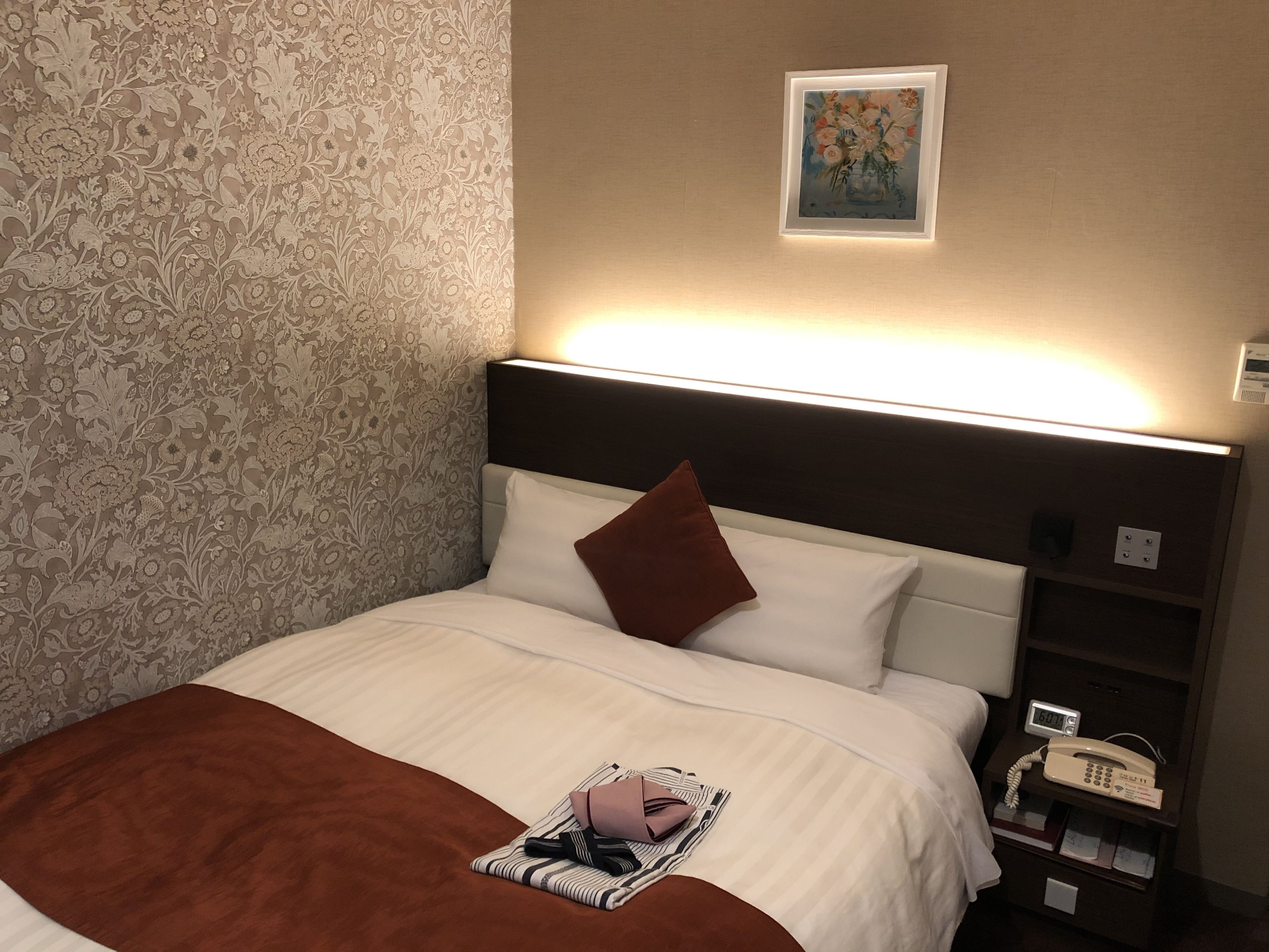 Cama bem arrumada com roupa de cama branca e castanha, um travesseiro castanho e uma toalha dobrada. Uma obra de arte floral emoldurada está na parede acima da cama.