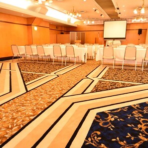 Sala de conferências com mesas, cadeiras, tela de projeção e piso coberto de carpete.