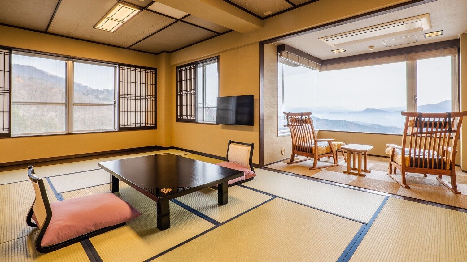Um quarto com tatames, uma mesa baixa de tatame, um banco acolchoado, uma TV e vista para montanhas através de grandes janelas.