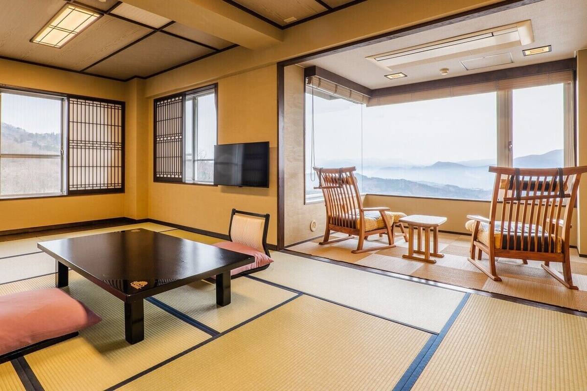 Um quarto com tatames, uma mesa baixa de tatame, um banco acolchoado, uma TV e vista para montanhas através de grandes janelas.