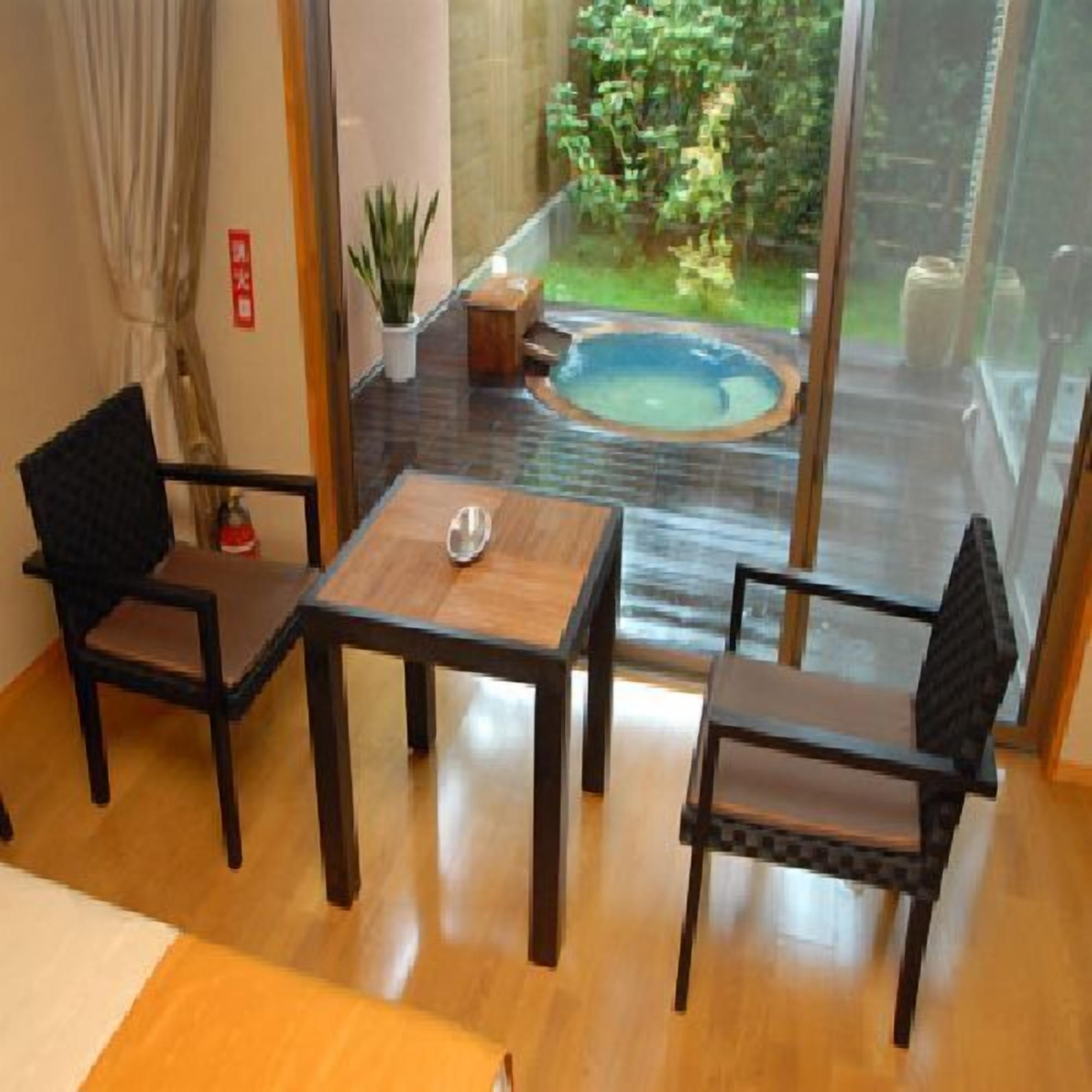 Quarto com porta de vidro que dá para um jardim com hidromassagem. Há duas cadeiras de madeira e uma pequena mesa com um copo.