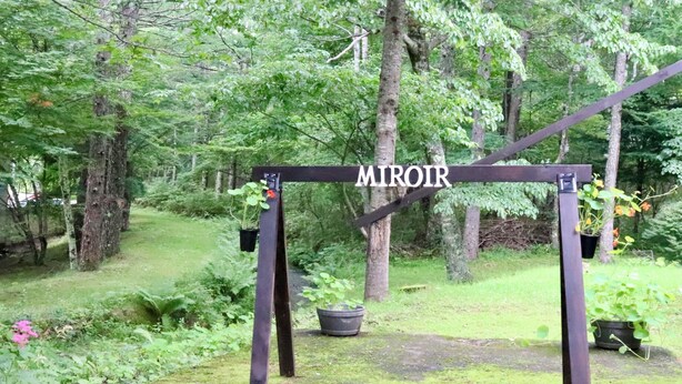 Uma placa de madeira com a palavra 'MIROIR' em letras brancas, rodeada por vegetação e árvores.