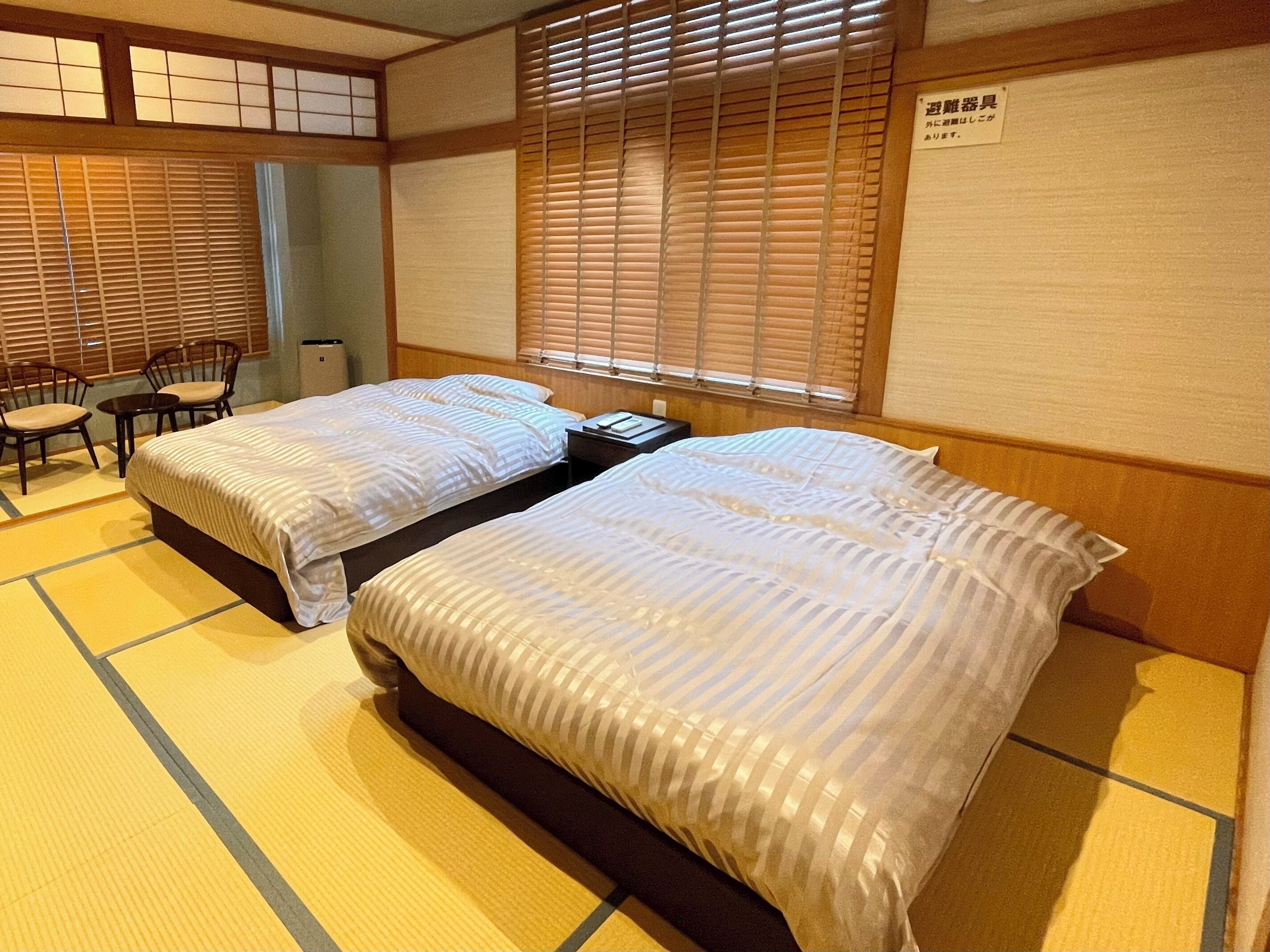 Um quarto tradicional em estilo japonês, com piso de tatame, duas camas com roupa de cama listrada, uma mesa pequena, cadeiras e portas deslizantes shoji.