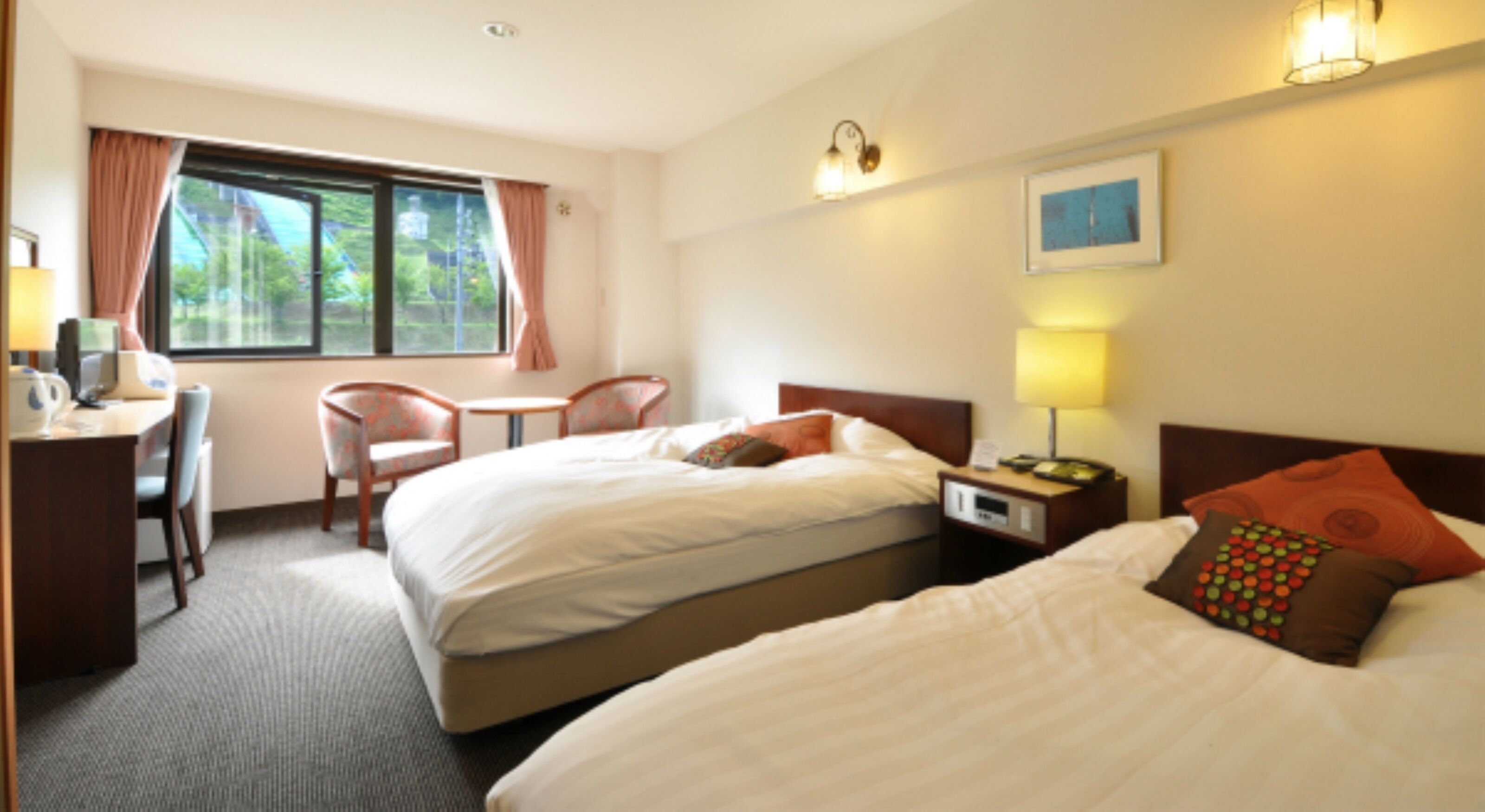 Quarto de hotel com duas camas, uma escrivaninha, uma cadeira, uma janela com cortinas e uma mesa de cabeceira com abajur.