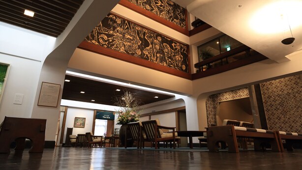 Um hall espaçoso com teto alto, vigas de madeira e uma grande parede decorativa. Há áreas de estar com cadeiras e mesas, e uma planta em vaso ao centro.