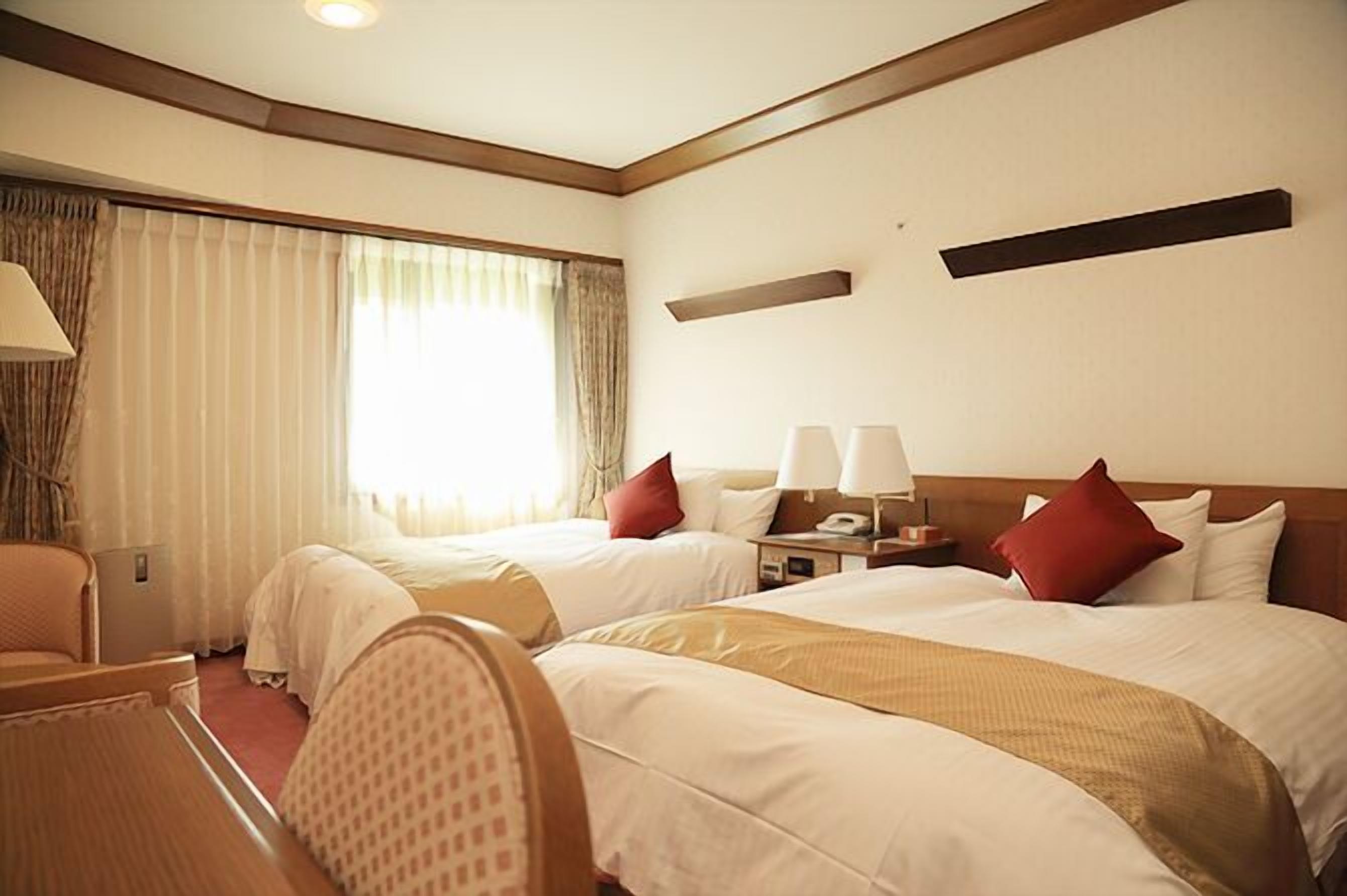 Quarto de hotel com duas camas, uma escrivaninha, uma cadeira e um abajur.