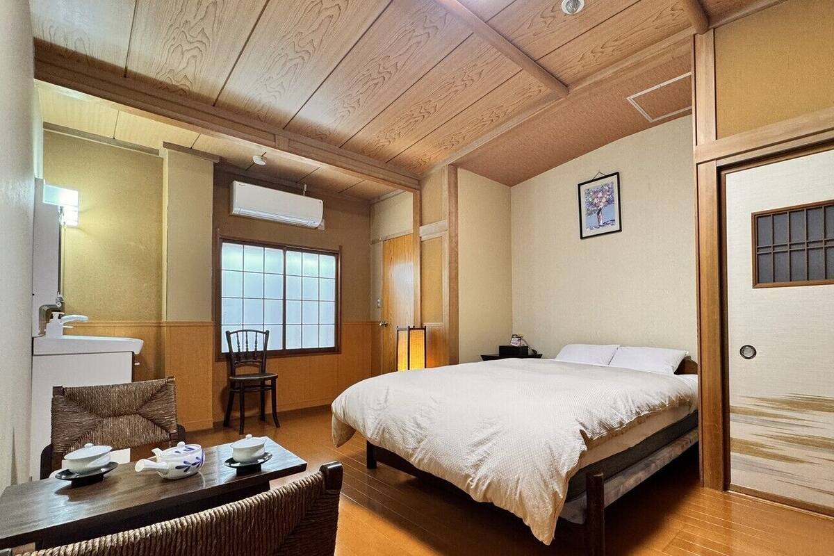 Quarto japonês tradicional com teto de madeira, uma cama, uma mesa de jantar com utensílios de chá e uma janela com vista.
