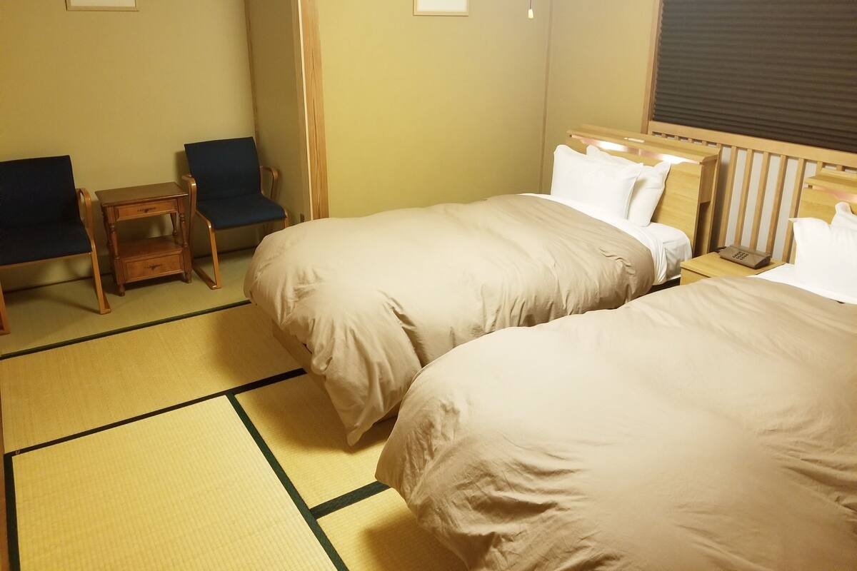 Quarto de hotel com duas camas, uma mesa pequena, uma cadeira e um futon.